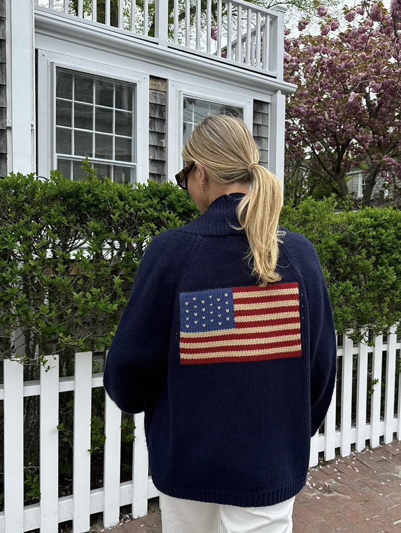 A woman with a ponytail stands facing a white picket fence and a house%2C wearing a navy sweater with an American flag pattern on the back. Blossoming trees add a spring vibe.